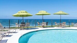 Uma área à beira da piscina com três guarda-sóis verdes, espreguiçadeiras e uma mesa com cadeiras.
