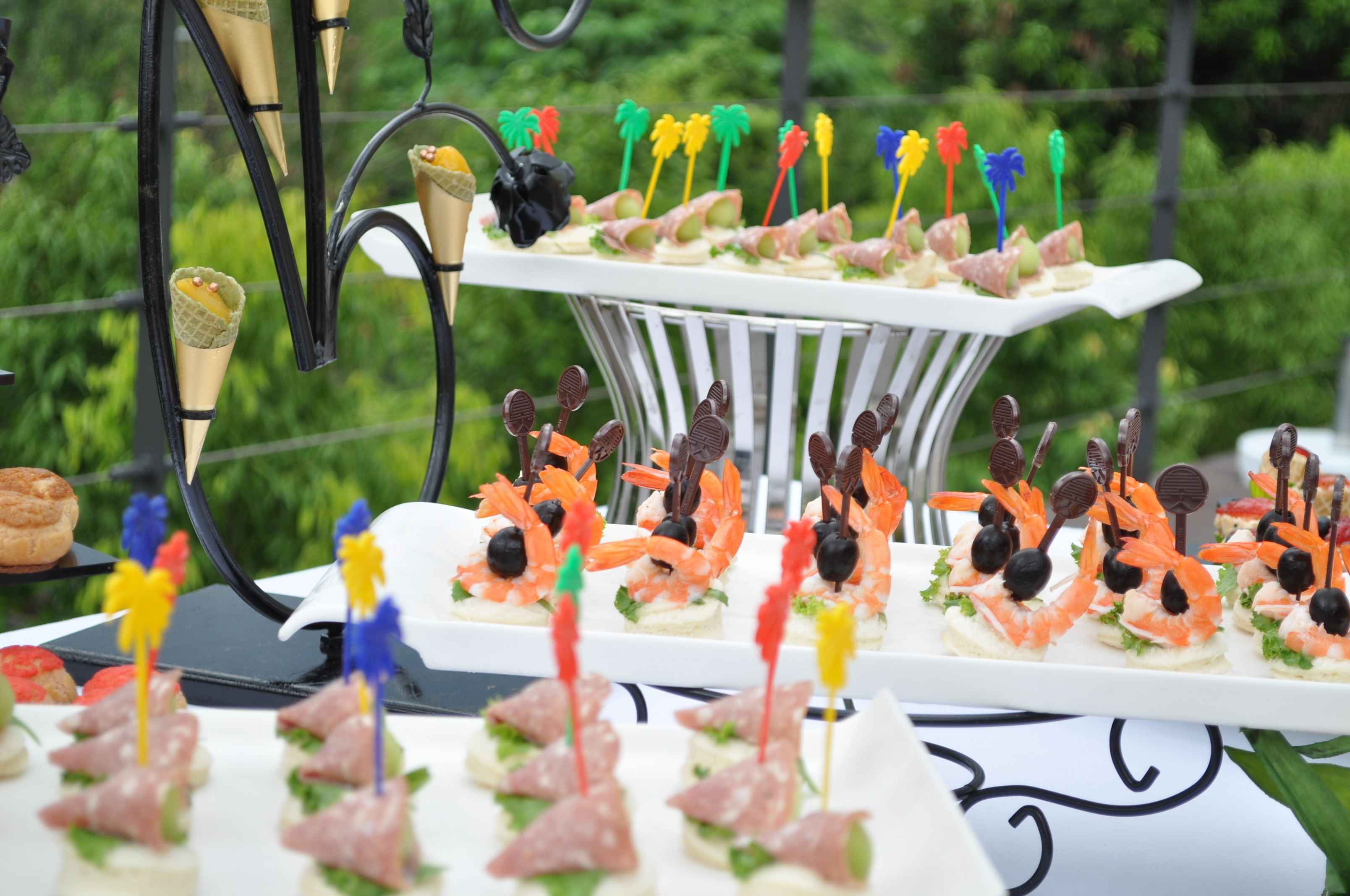 Uma mesa de buffet com diversos aperitivos, incluindo espetos de camarão e carne, dispostos em pratos brancos.