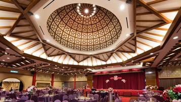 Uma sala de banquetes com mesas redondas, cadeiras roxas e um palco com cortinas vermelhas.