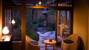 Um quarto tradicional japonês com tatames, uma mesa baixa, duas cadeiras de tatame e vista para um jardim com pedras e um lampião de pedra.