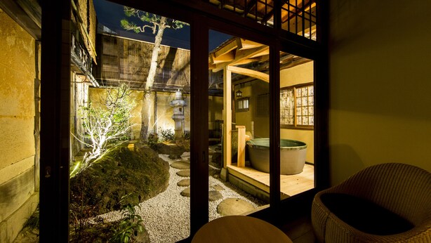 Um quarto tradicional japonês com piso de tatame, um candeeiro de pedra e uma grande porta de madeira que dá para um jardim exterior com um lago de carpas e pedras para caminhar.
