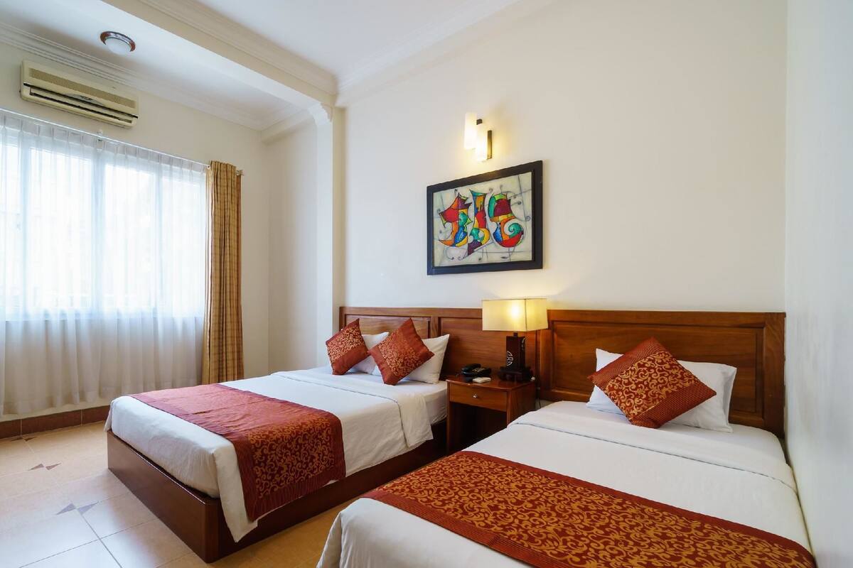 Quarto de hotel com duas camas, cada uma com cabeceira de madeira, roupa de cama branca e mantas decorativas estampadas em vermelho. Há uma mesa de cabeceira com uma luminária, ar-condicionado fixado na parede e uma obra de arte emoldurada acima das camas.