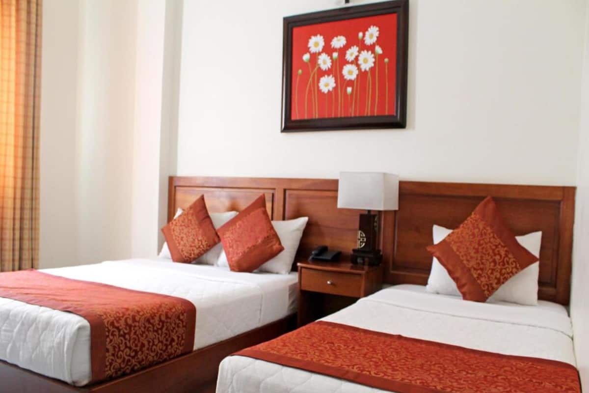 Quarto de hotel com duas camas, cada uma com cabeceira de madeira, roupa de cama branca e almofadas decorativas vermelhas. Um quadro com flores está pendurado na parede acima das camas.
