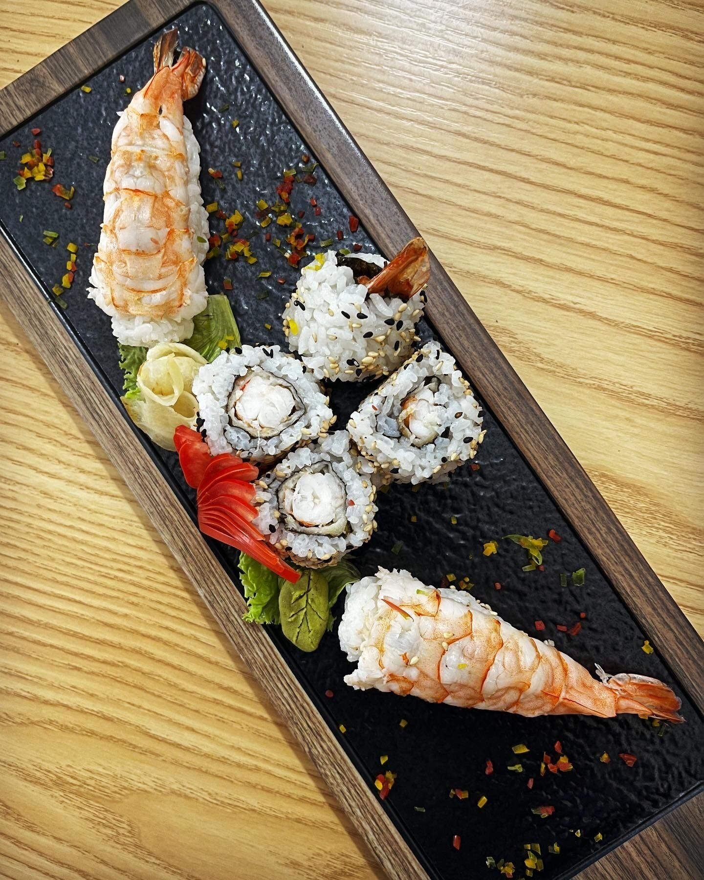 Uma bandeja de sushi com diversos rolinhos e camarões em um prato de ardósia preta.