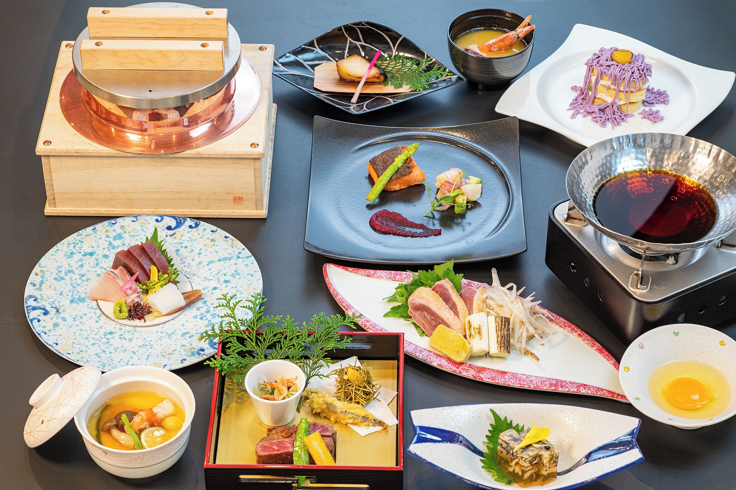 Diversos pratos japoneses dispostos sobre uma mesa, incluindo sushi, sashimi e outras iguarias, com um bule e xícaras à vista.