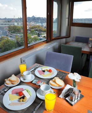 Uma mesa de café da manhã com pratos de comida, um copo de suco de laranja e vista para a cidade através de grandes janelas.