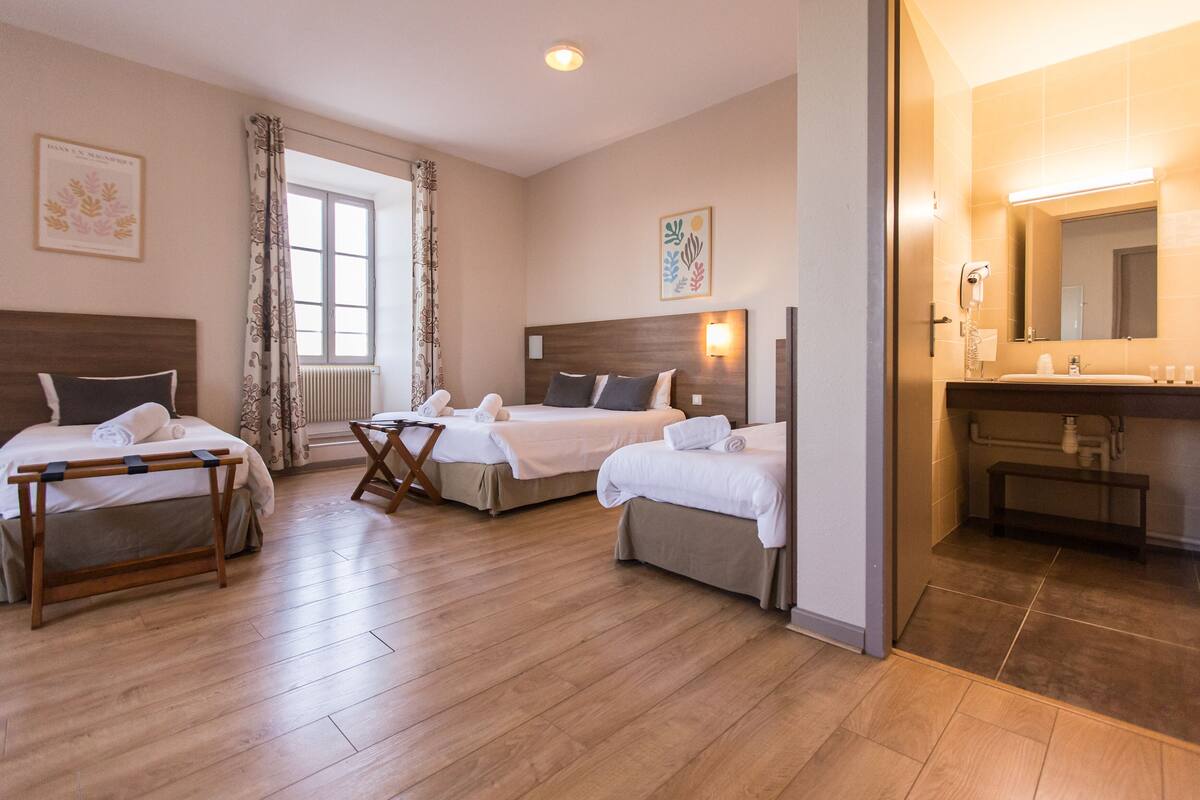 Quarto de hotel com três camas, piso de madeira, janela com cortinas, banheiro com pia e espelho, e obras de arte nas paredes.