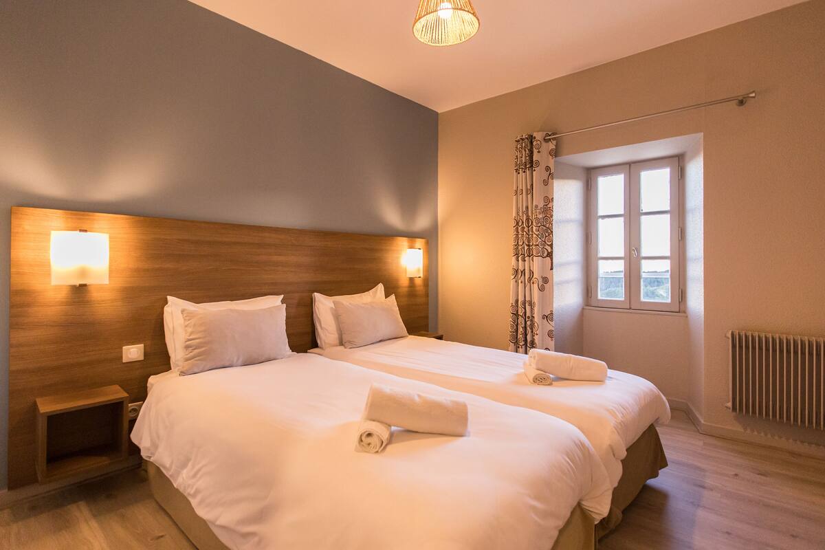 Uma cama de casal com lençóis brancos, cabeceira de madeira, abajures e uma janela com cortinas.
