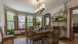 Uma sala de jantar com uma mesa de madeira posta para seis pessoas, rodeada por seis cadeiras. Há um candelabro sobre a mesa e um vaso de plantas no parapeito da janela.