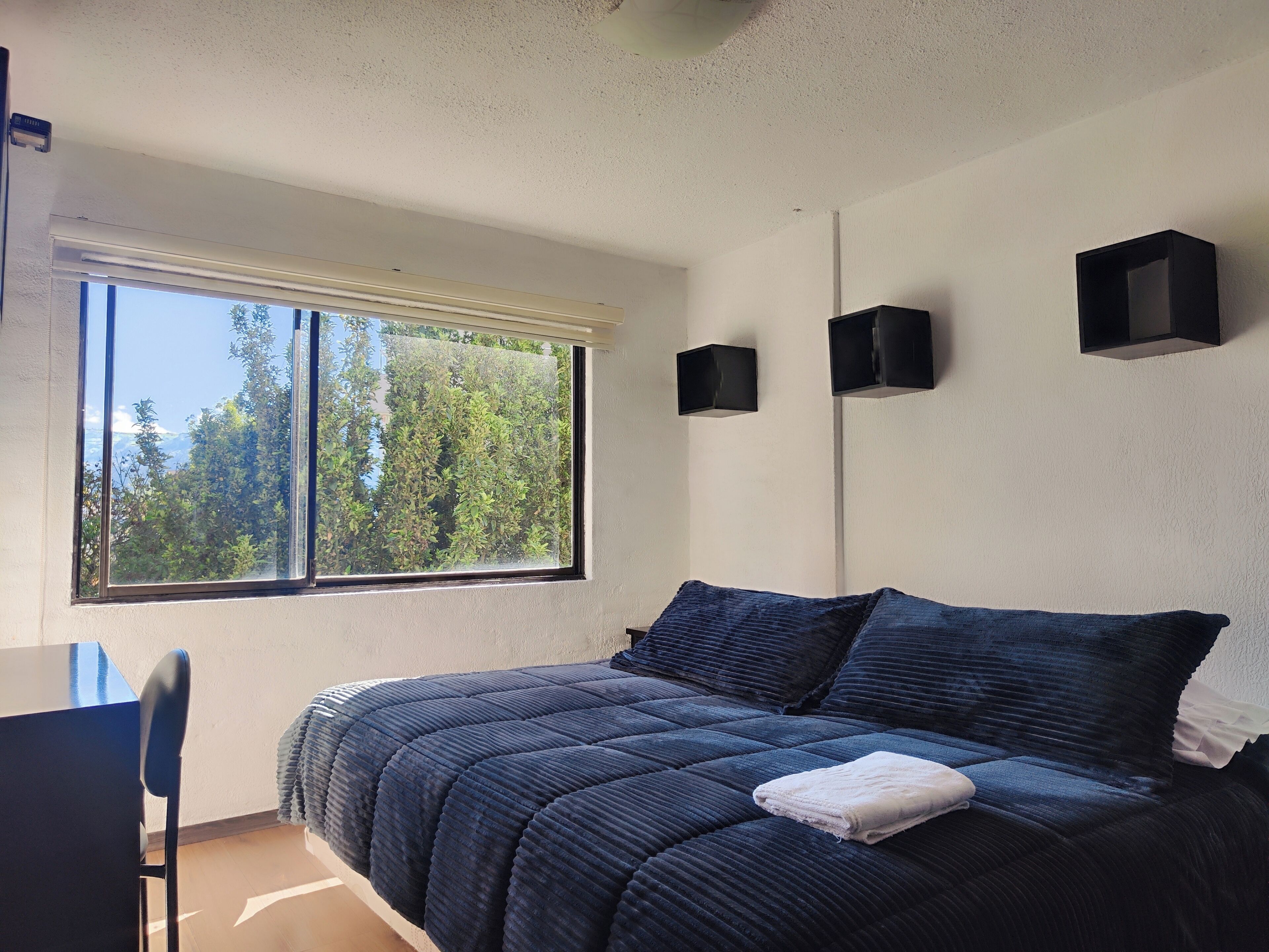 Um quarto com cama, escrivaninha e janela com vista para árvores.