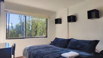 Um quarto com cama, escrivaninha e janela com vista para árvores.