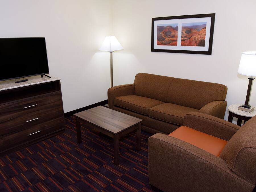 Quarto de hotel com televisão de tela plana sobre um móvel de madeira, um sofá marrom, uma poltrona laranja, uma pequena mesa de centro de madeira e duas luminárias de chão.