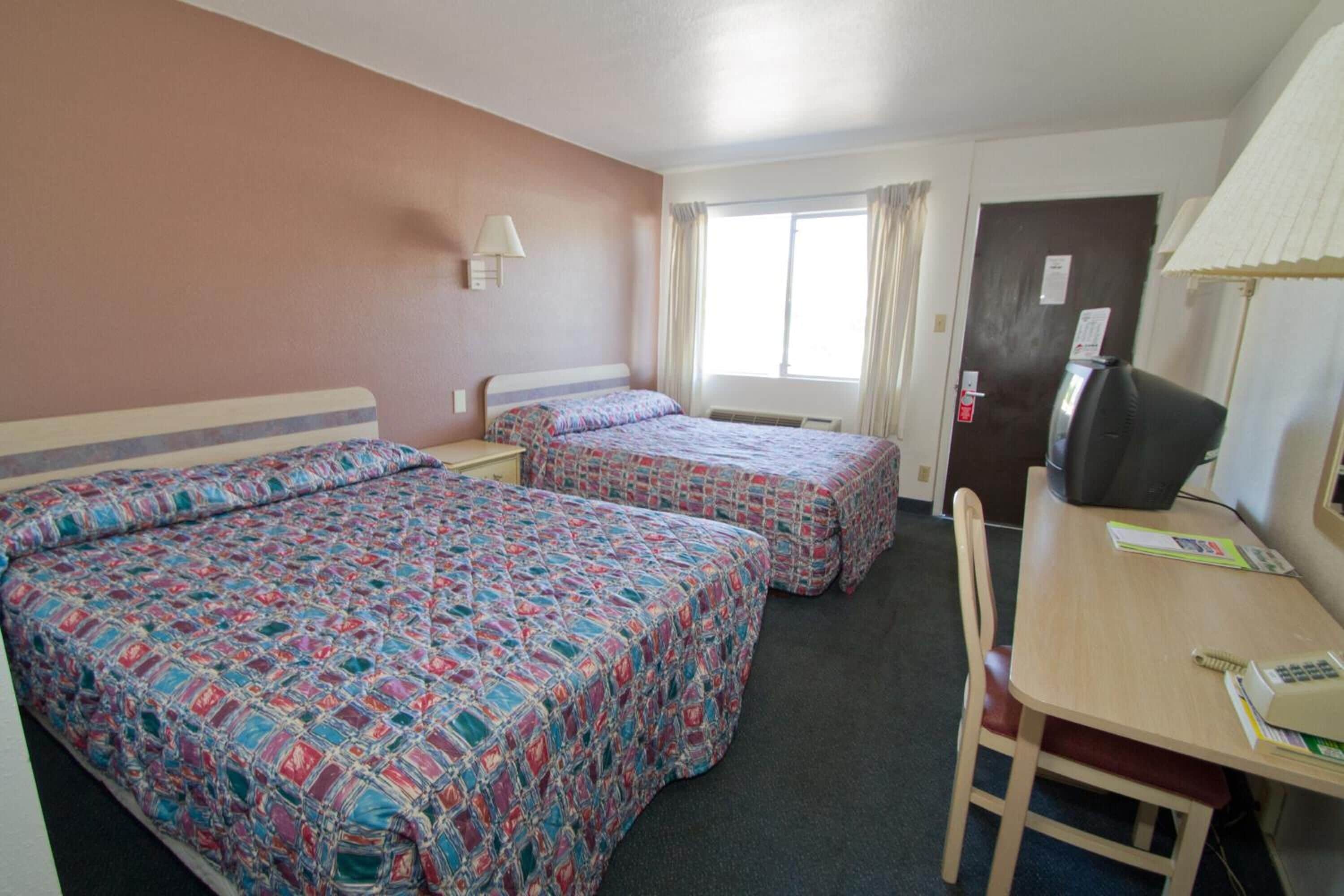 Quarto de hotel com duas camas, uma escrivaninha, uma cadeira, um abajur e uma janela.
