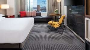 Quarto de hotel com uma cama grande, um sofá, uma mesinha com um vaso, uma televisão e vista para a cidade.