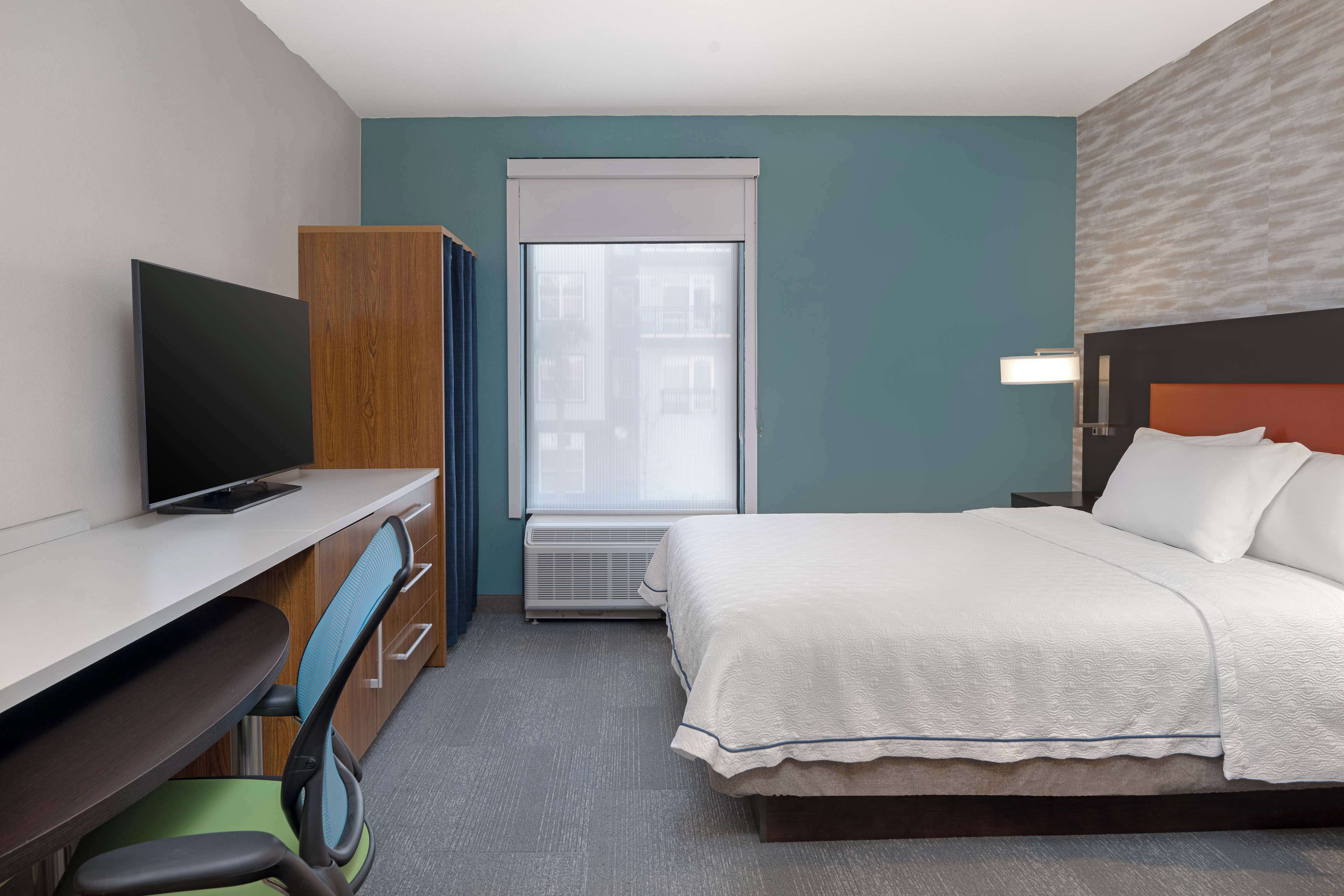 Quarto de hotel com cama, escrivaninha com televisão, cadeira, guarda-roupa e janela com persianas.