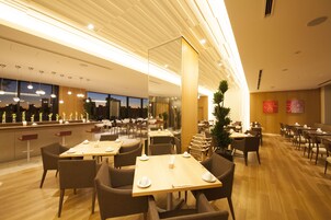 Um restaurante moderno com mesas e cadeiras de madeira, área de bar e vista para a cidade através de grandes janelas.