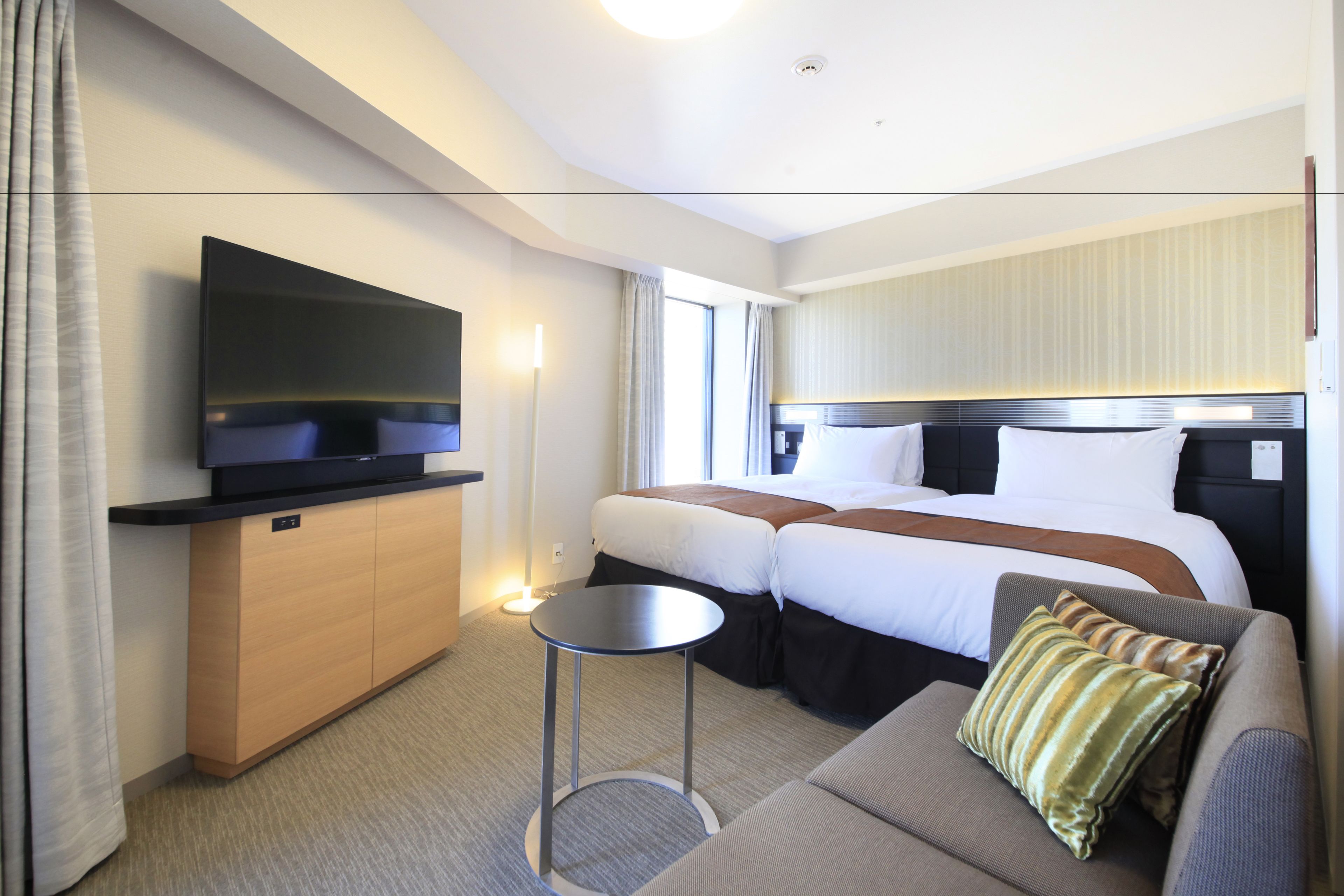 Quarto de hotel com cama, sofá, televisão em móvel, mesinha e janela com cortinas.