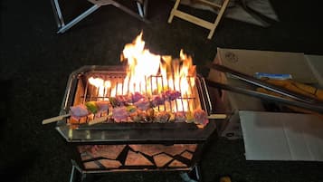 Uma churrasqueira portátil com espetos de carne e legumes, cercada por cadeiras de camping e uma tenda.