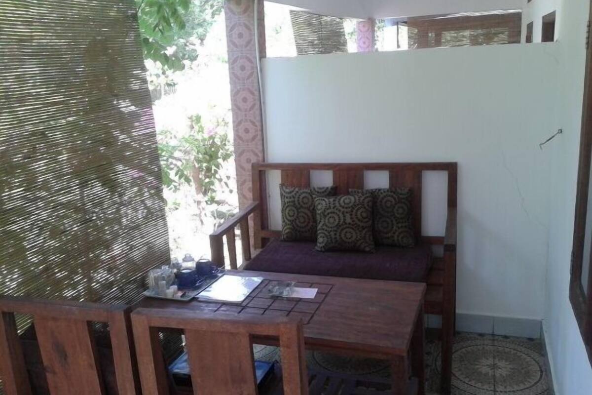 Uma pequena área externa coberta com mesa e cadeiras de madeira, um banco acolchoado com almofadas estampadas e vista para o verde através de uma janela decorada.