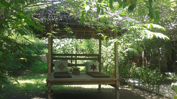 Um banco de madeira com almofadas sob uma estrutura de telhado de palha, rodeado por vegetação exuberante.