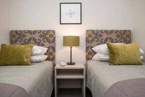 Quarto duplo com duas camas, cada uma com cabeceira estampada, mesa de cabeceira com abajur e relógio.
