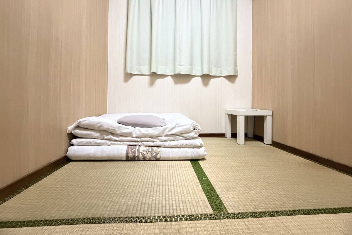 Quarto com piso de tatame, cama com almofadas, mesa pequena e janela com cortina.
