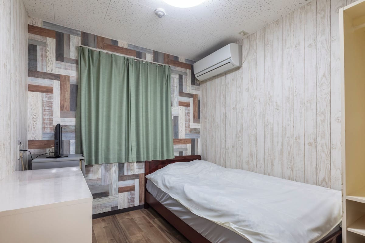 Quarto pequeno, com cama de casal, cabeceira de madeira, cortina verde e unidade de ar condicionado branca fixada na parede.