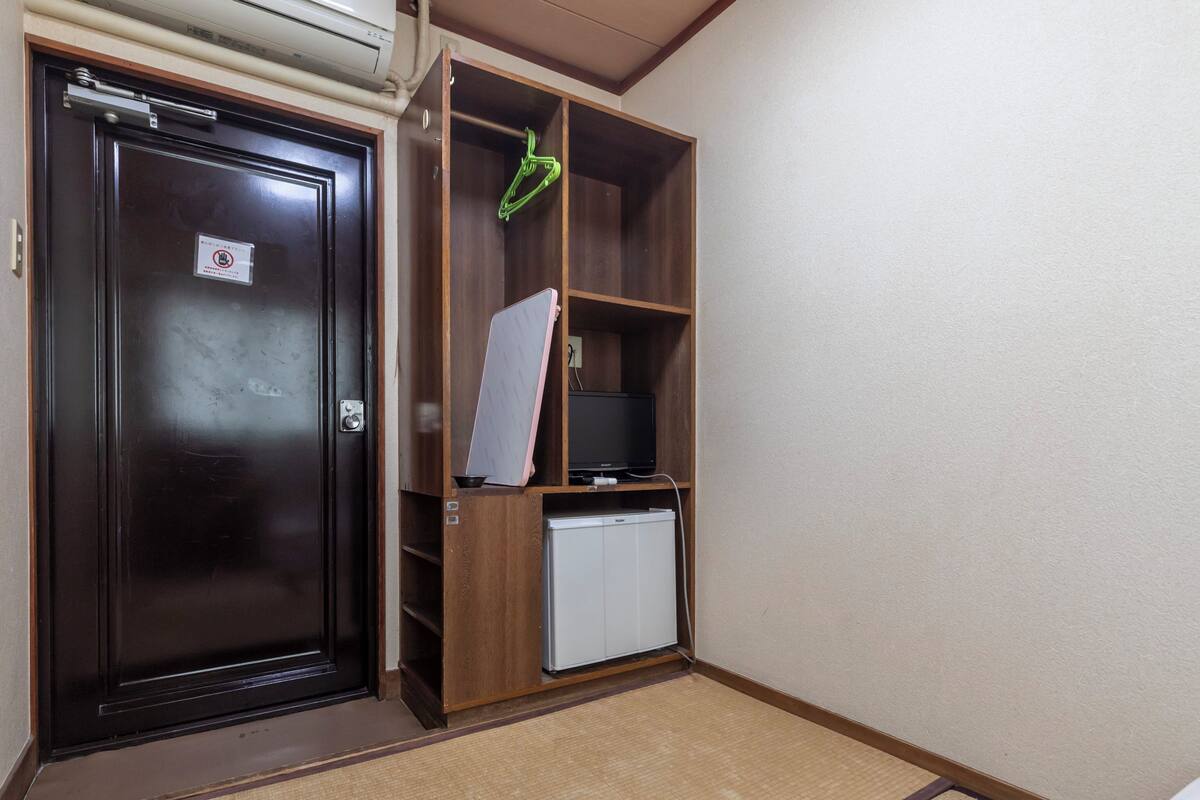 Quarto de hotel com porta de madeira escura, um pequeno frigorífico branco, uma televisão sobre uma prateleira de madeira e um cabide verde.
