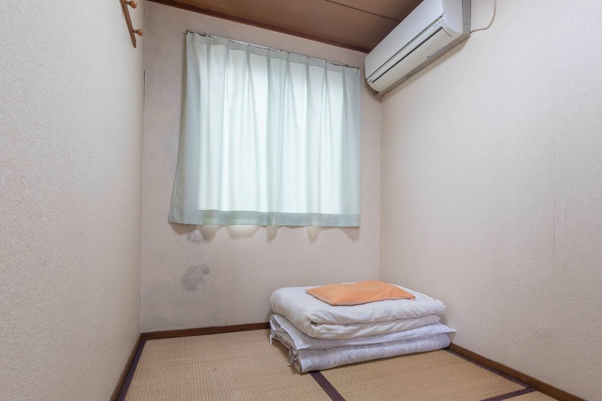 Quarto pequeno e limpo com futon, janela com cortinas leves e ar condicionado na parede.