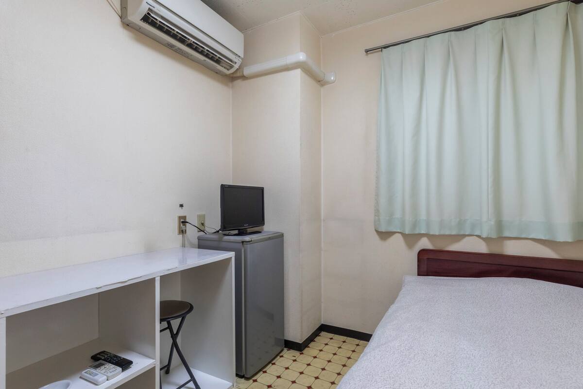 Um quarto pequeno e simples, com uma cama, uma escrivaninha com computador, um banquinho, uma prateleira com uma tigela e um controle remoto, e uma janela com cortina.