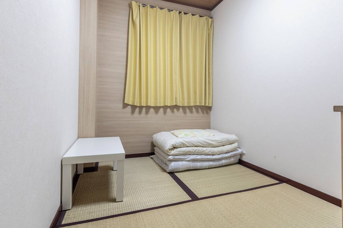 Quarto pequeno e limpo com chão de esteira de tatame, uma mesa branca e uma pilha de roupa de cama dobrada na parede.