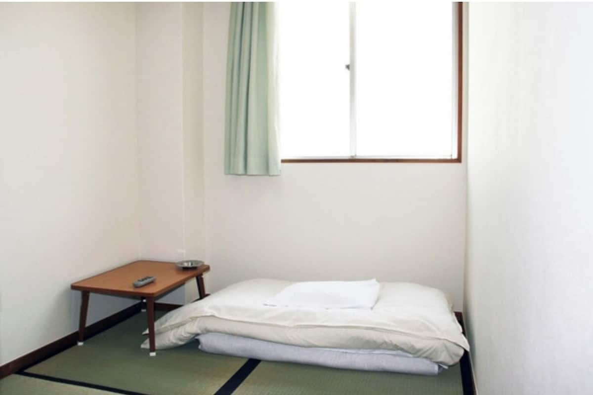 Quarto pequeno e modesto, com uma cama de solteiro, uma mesinha e uma janela.