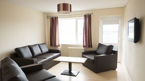Uma sala de estar moderna com um sofá seccional, uma mesa de centro e duas poltronas. Há uma janela grande com cortinas, uma televisão de tela plana na parede e um lustre.