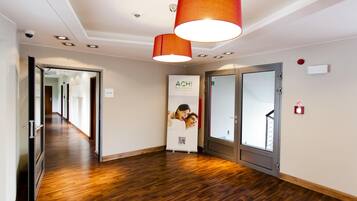 Corredor de hotel com piso de madeira, área de recepção com um banner e uma porta com painel de vidro.