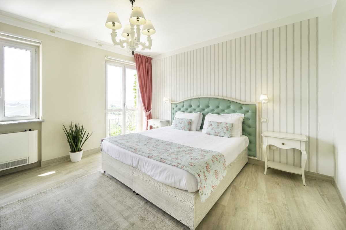 Quarto com uma cama grande, um candelabro, uma planta em vaso e uma janela com cortinas.