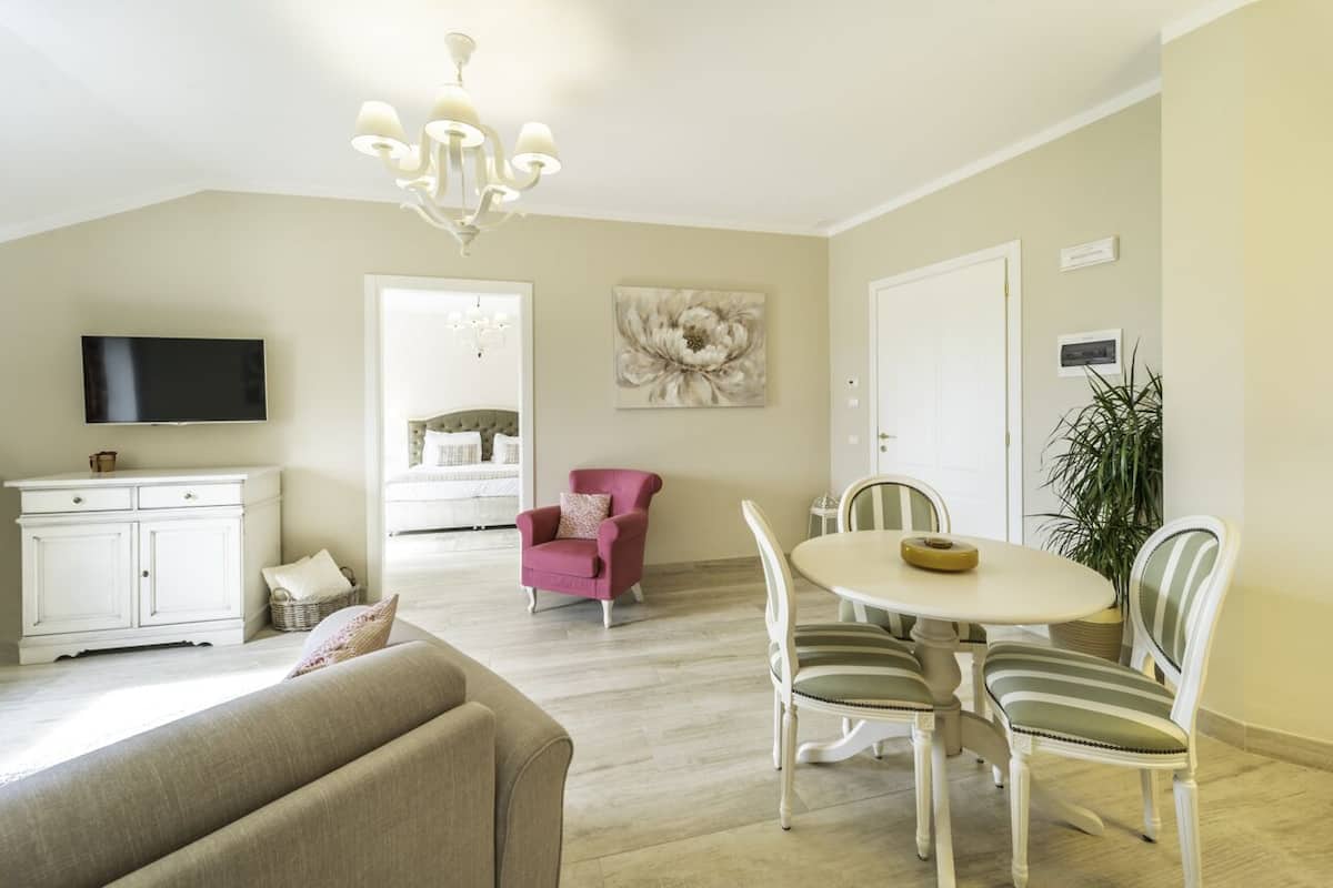 Uma sala de estar espaçosa com um sofá, uma mesa de jantar com cadeiras, uma poltrona rosa, um lustre e uma televisão na parede.