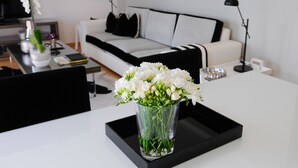 Uma sala de estar moderna com um sofá branco, almofadas preto e branco, um vaso de vidro com flores brancas sobre uma bandeja preta e uma televisão ao fundo.