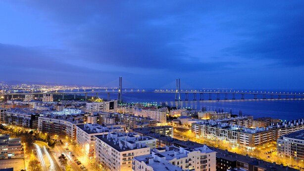 Vista urbana ao entardecer, com uma ponte imponente ao fundo e a orla marítima à mostra.