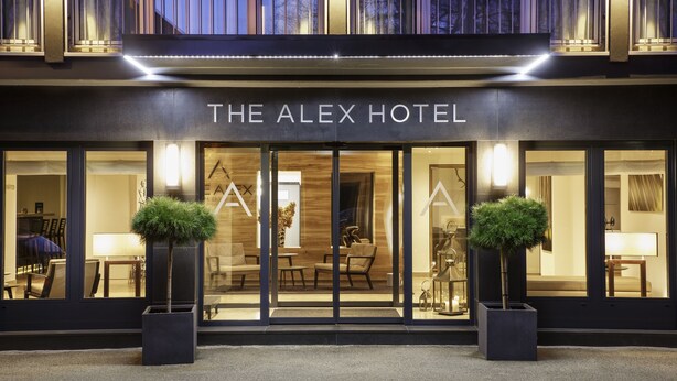 O Hotel Alex, com fachada moderna, grandes janelas de vidro e dois vasos de plantas em frente.