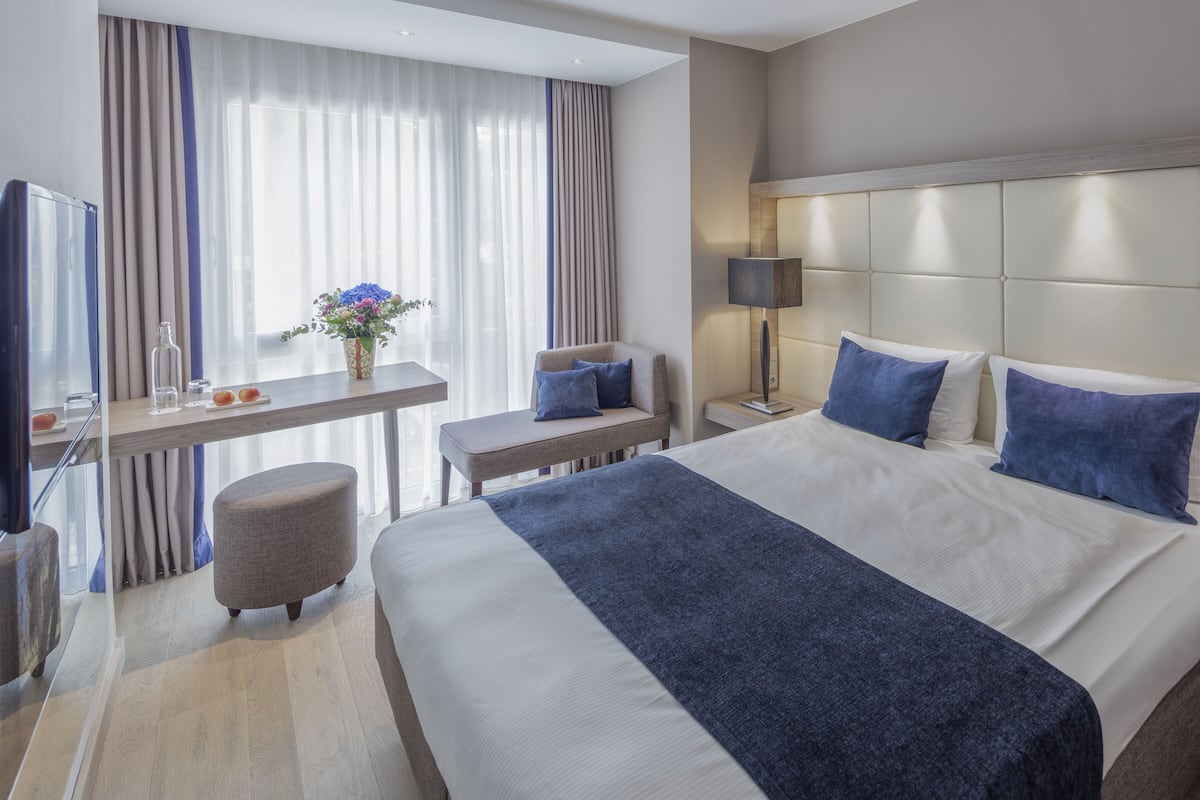 Quarto de hotel moderno com uma cama grande, uma escrivaninha, uma cadeira e uma pequena mesa com um vaso de flores.