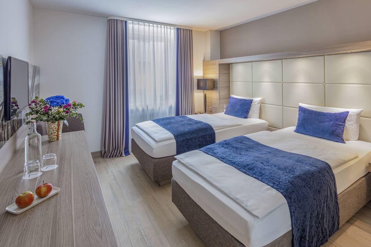 Quarto de hotel com duas camas, uma escrivaninha com um vaso de flores e uma janela grande com cortinas.