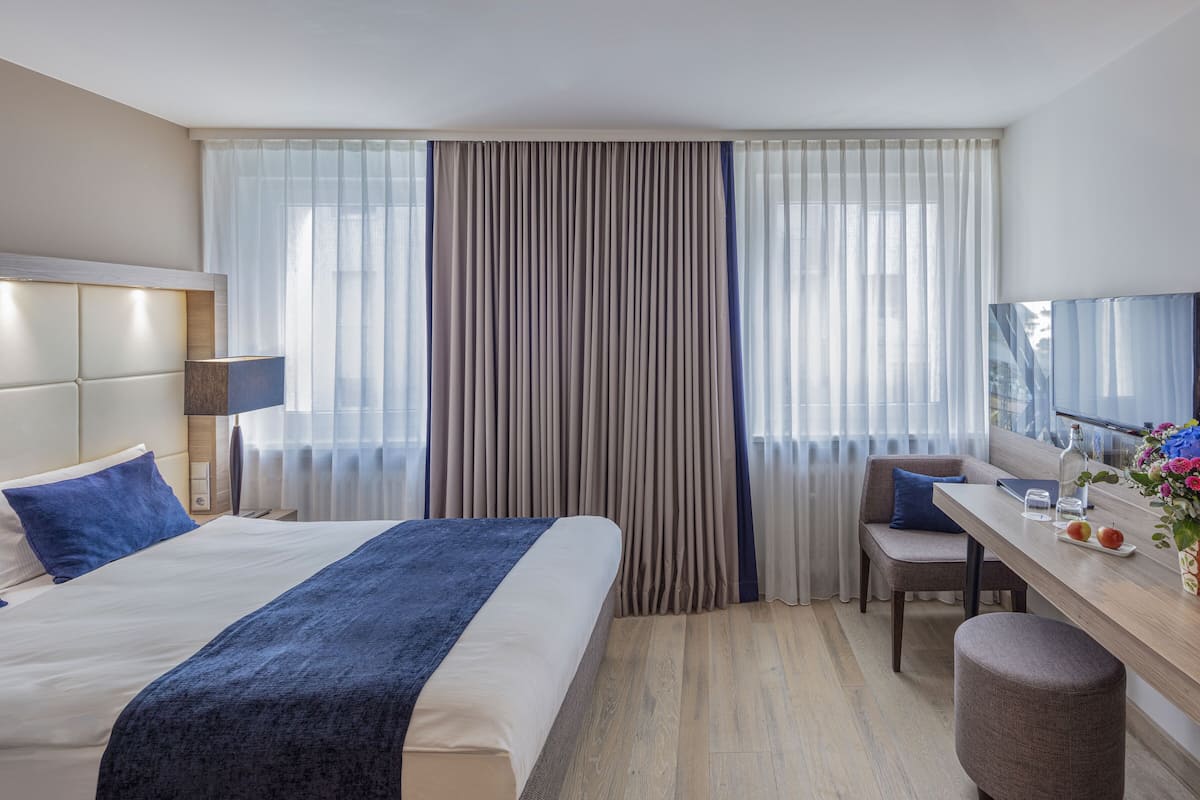 Quarto de hotel com uma cama grande, uma escrivaninha com cadeira, uma televisão e uma janela com cortinas.