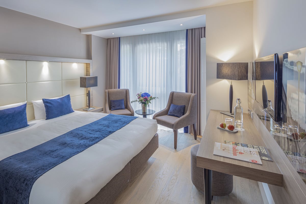 Quarto de hotel com uma cama grande, duas poltronas, uma mesinha com um vaso de flores, um tabuleiro com maçãs e uma garrafa, e uma revista sobre a mesa.