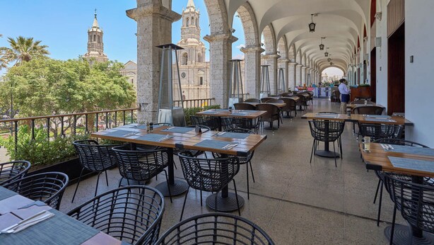Uma área de refeições ao ar livre com mesas e cadeiras, vista para uma igreja com um campanário, e um céu azul claro.