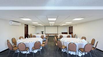 Uma sala de conferências com mesas redondas, cadeiras, uma tela de projetor e um quadro branco.