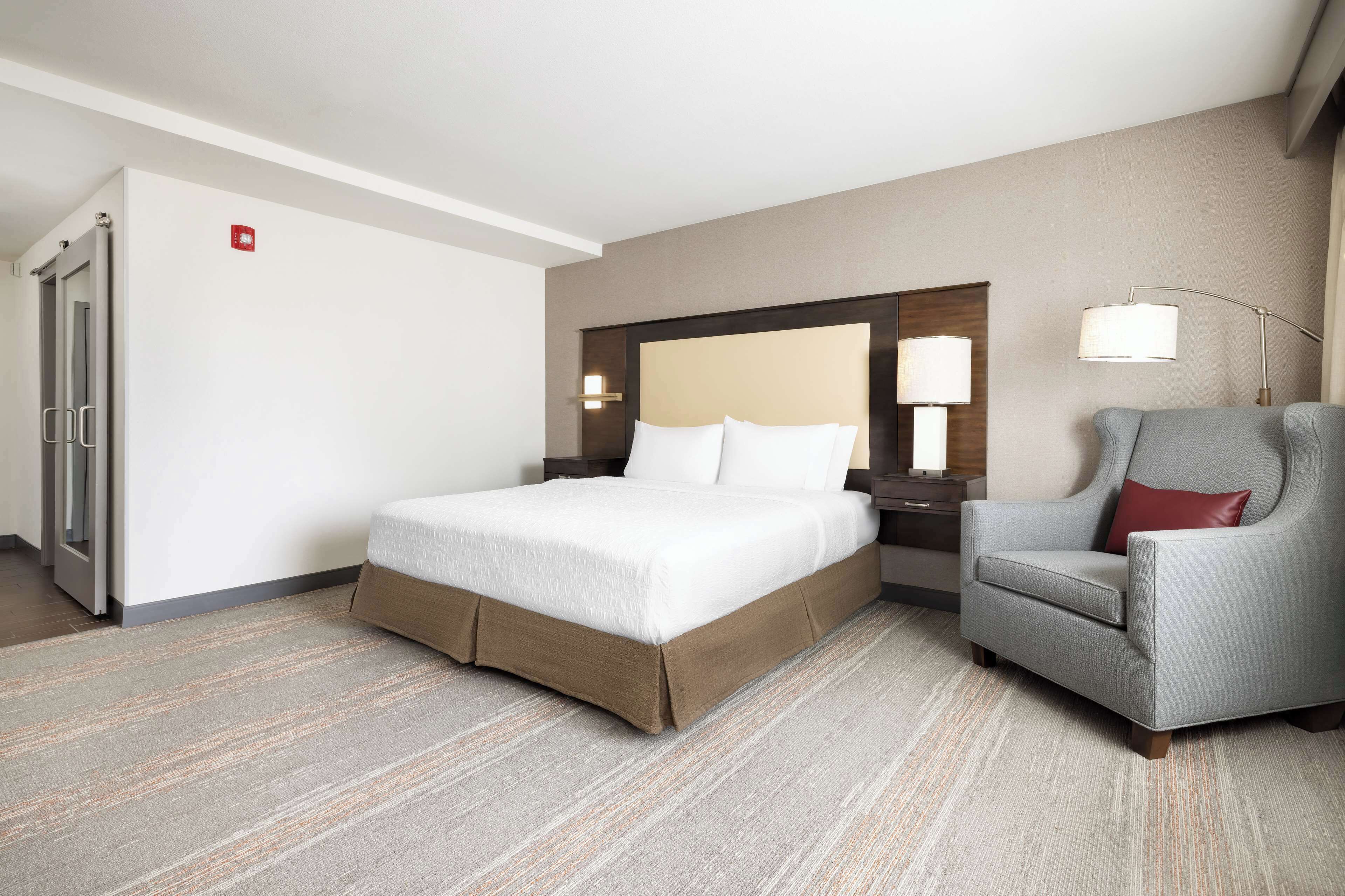Quarto de hotel com uma cama, uma cadeira, uma mesinha de cabeceira e um abajur.