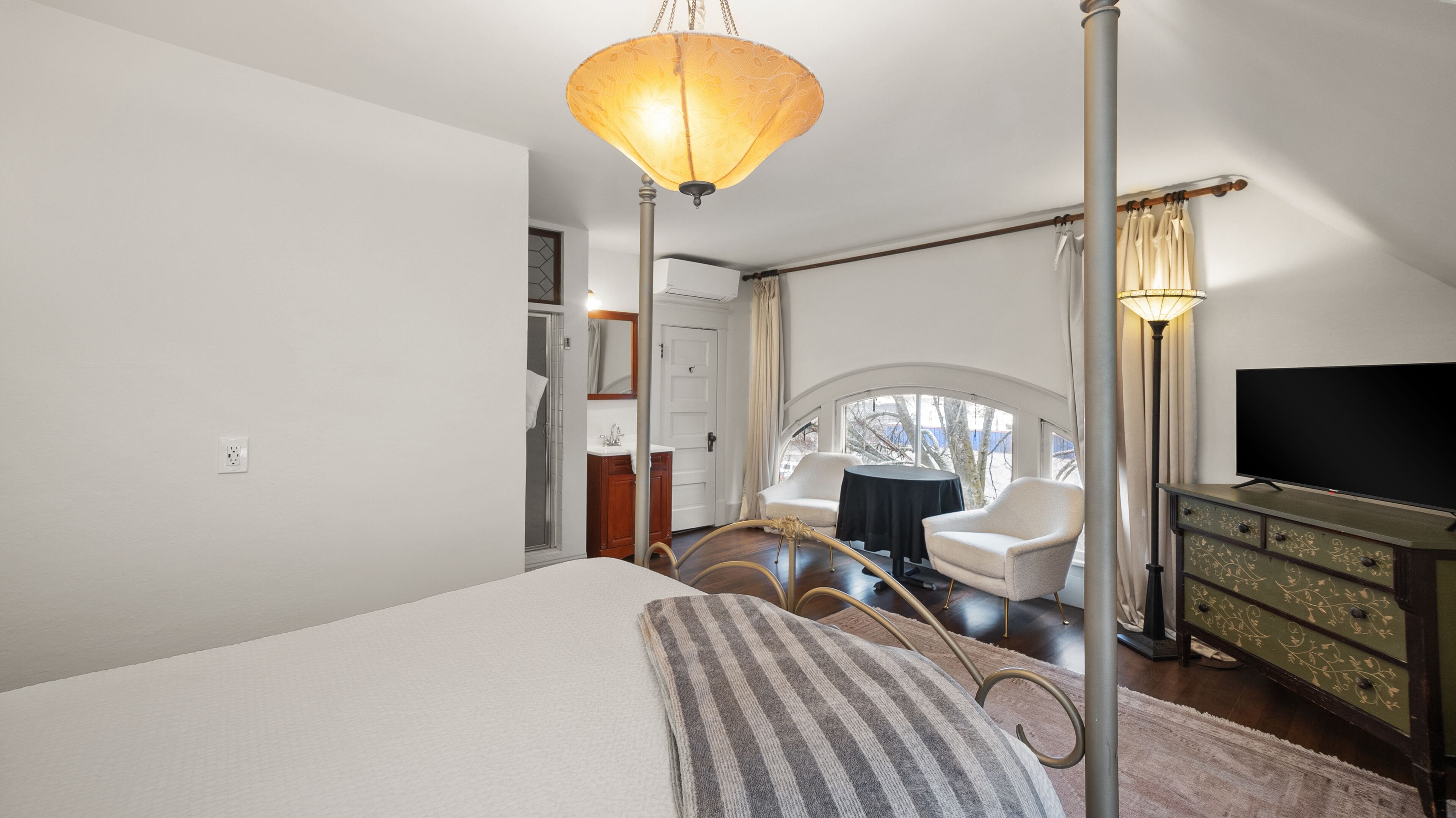 Quarto com cama, televisão, lustre, penteadeira e vista para o exterior.