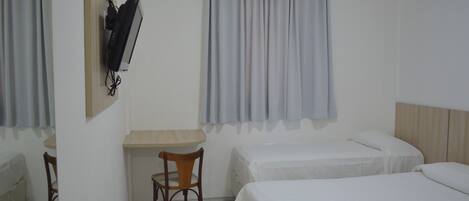 Quarto de hotel com uma cama, uma escrivaninha, uma cadeira e uma TV fixada na parede.