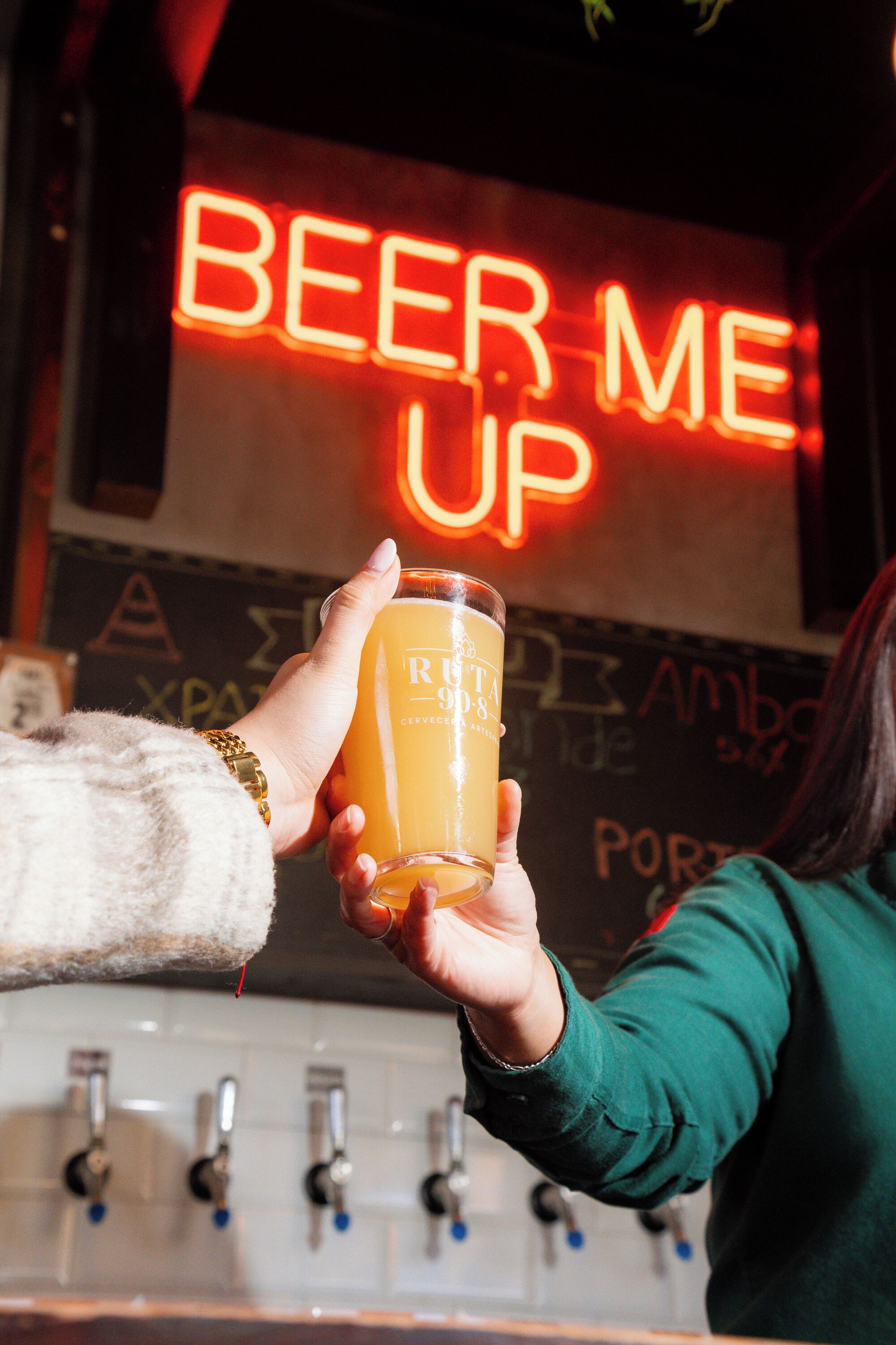 Uma pessoa segurando um copo de cerveja com um letreiro de neon ao fundo que diz 'BEER ME UP'.
