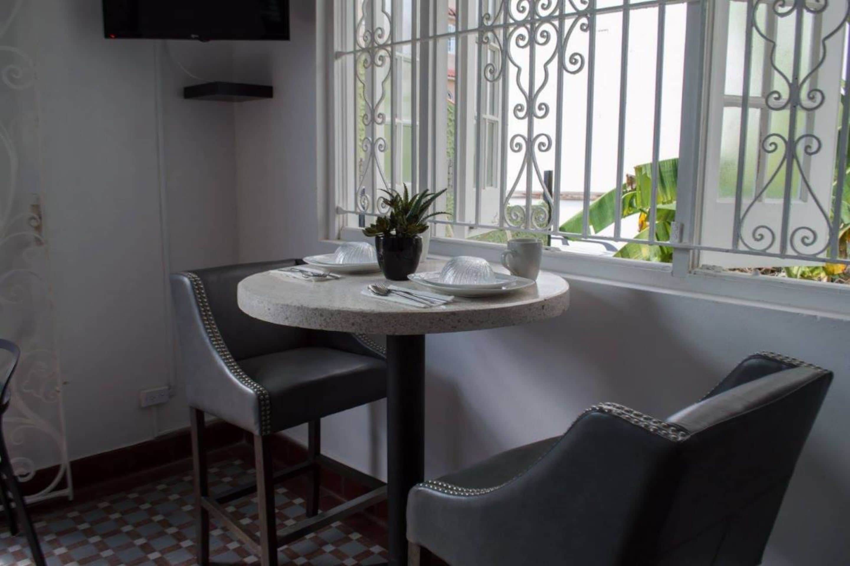 Uma área de refeições com uma mesa redonda, duas cadeiras, um vaso de planta e uma janela com grades decorativas.